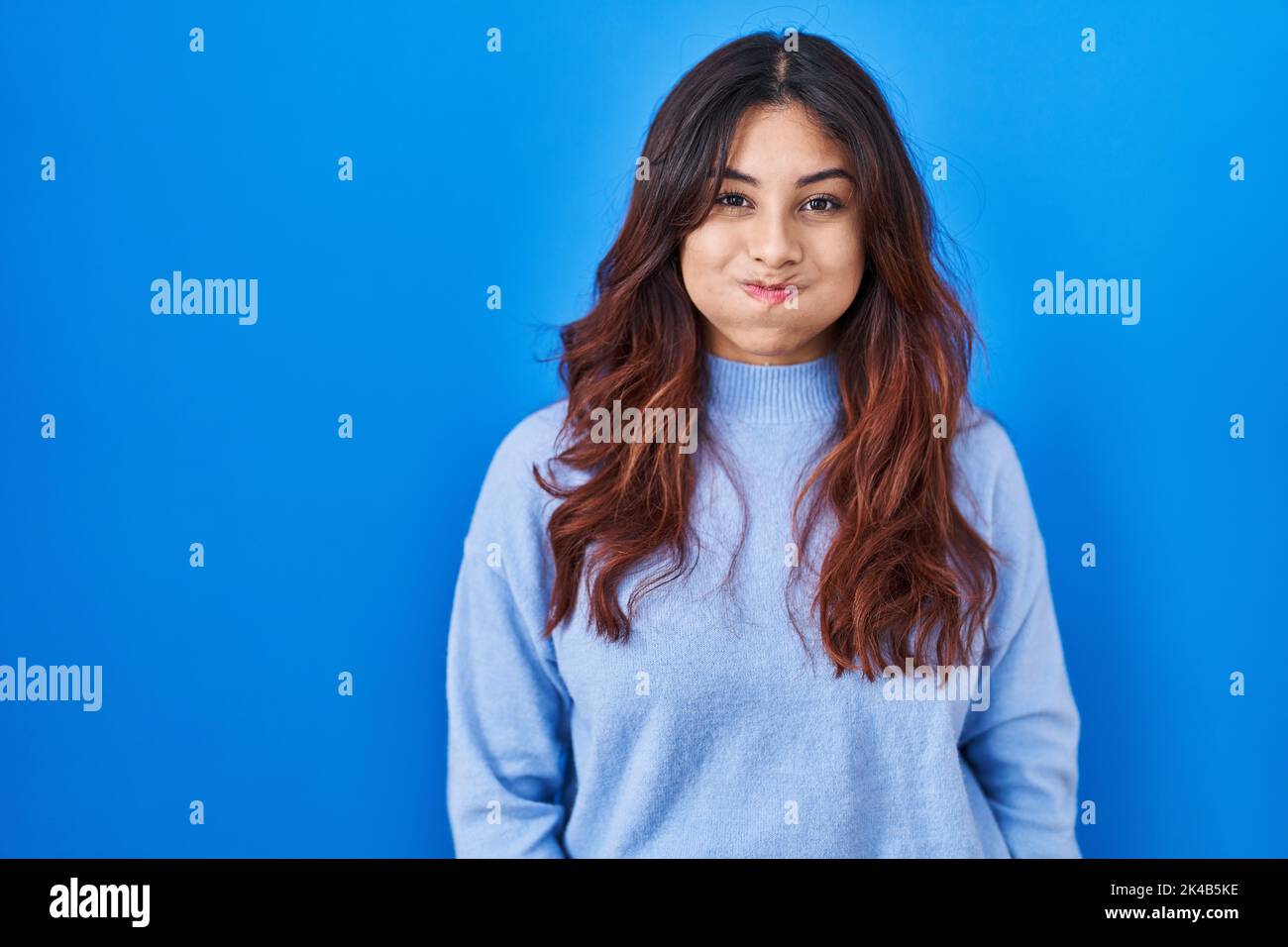 Hispanic young woman standing over blue background puffing cheeks with ...