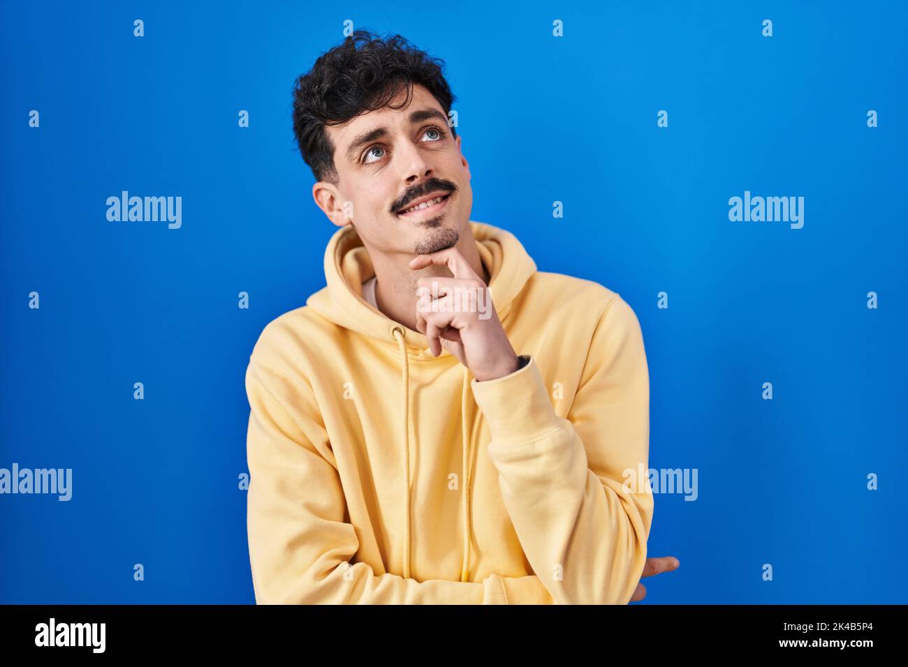 Hispanic man standing over blue background with hand on chin thinking ...