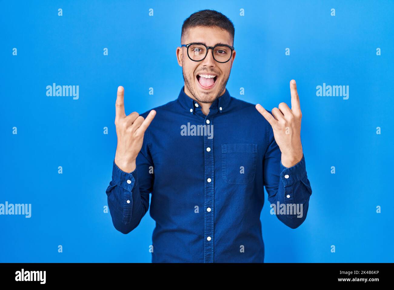 Young hispanic man wearing glasses over blue background shouting with ...