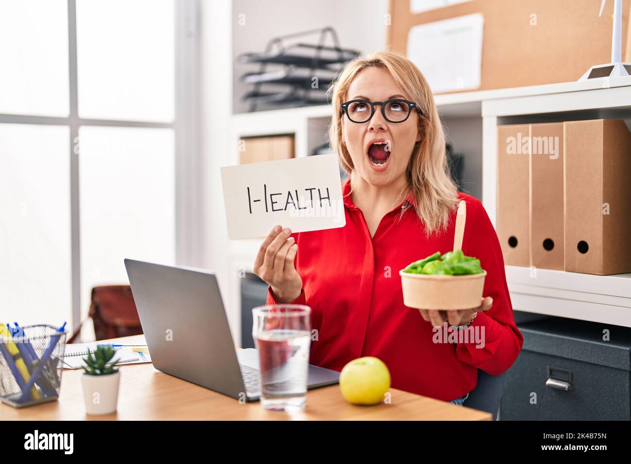 Blonde woman working at the office eating healthy food angry and mad ...