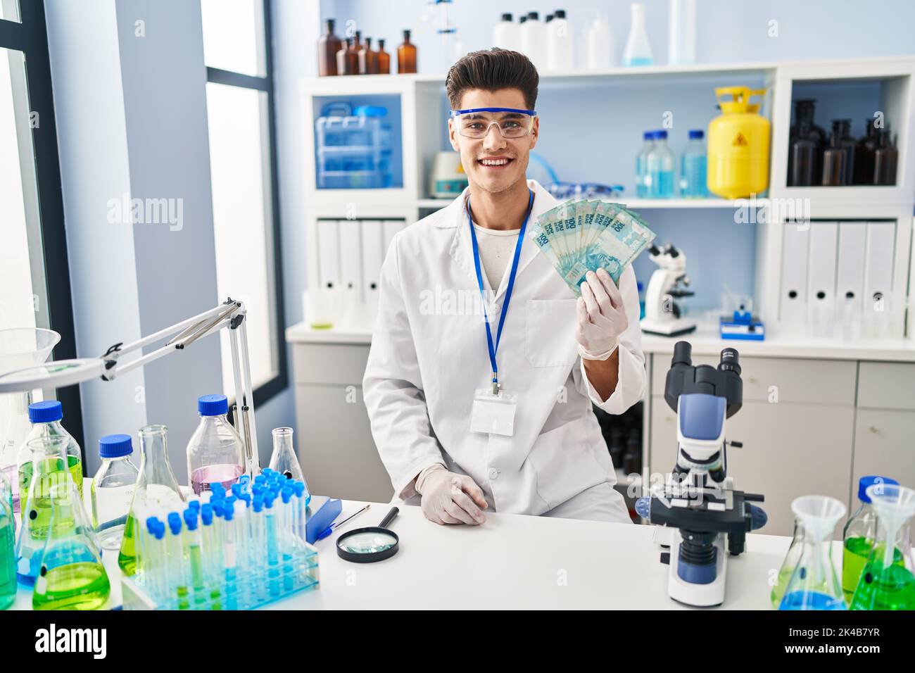 Young hispanic man working at scientist laboratory holding brazilian reals looking positive and ...