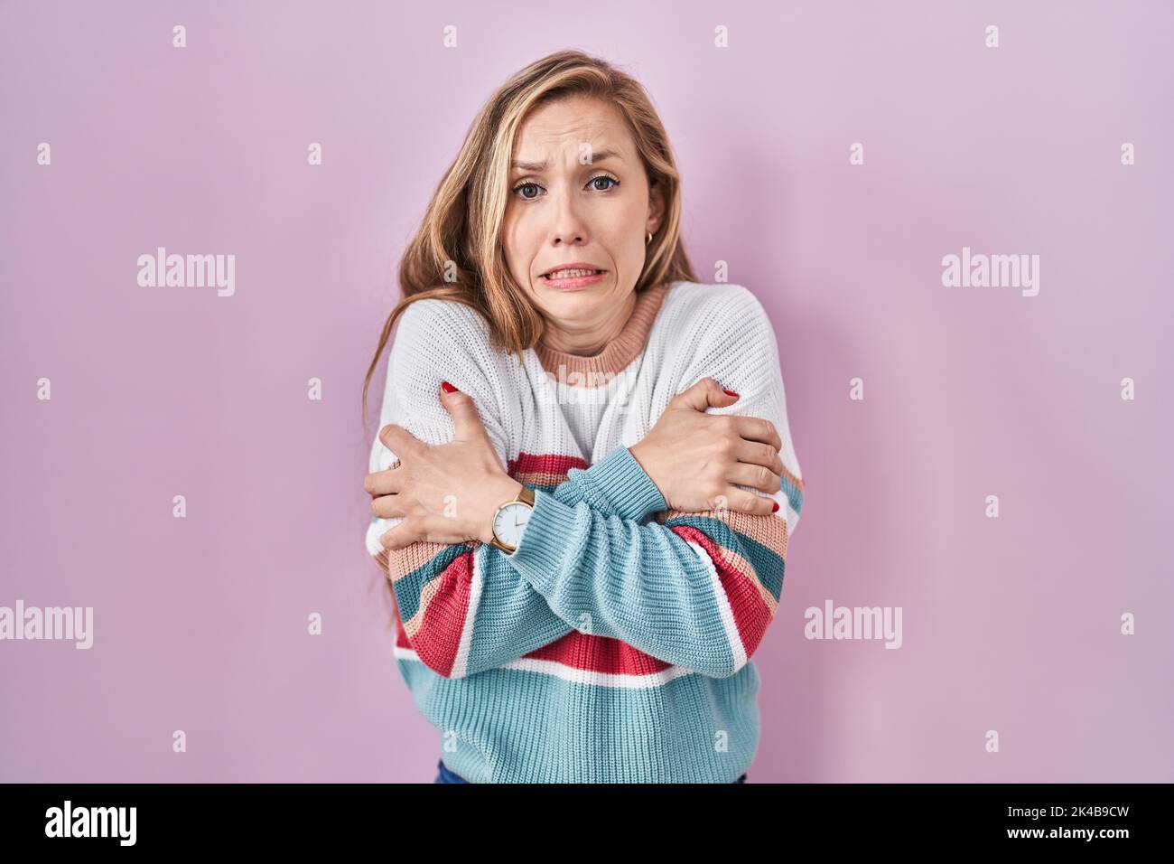 Young blonde woman standing over pink background shaking and freezing ...