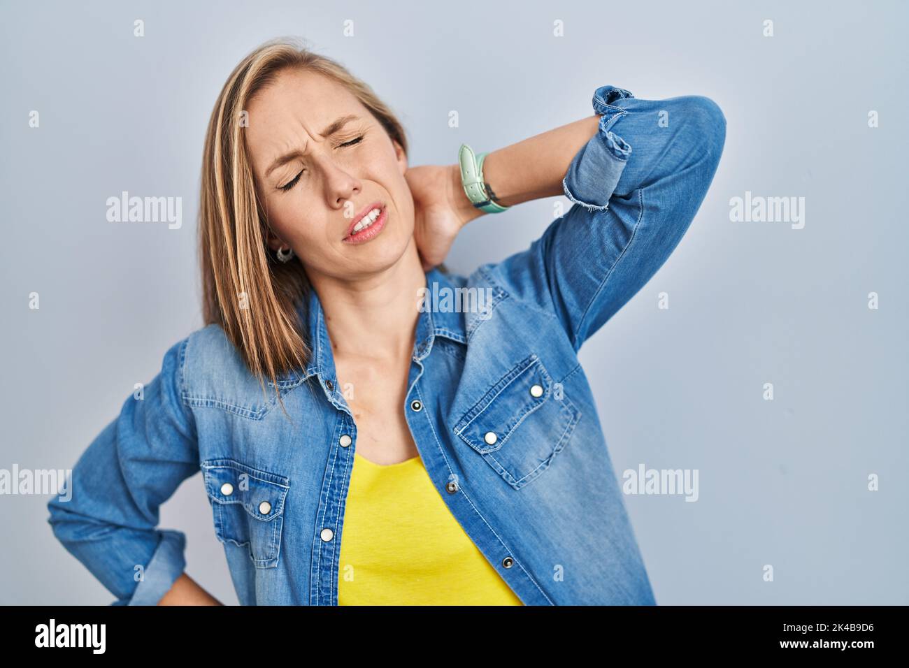 Young blonde woman standing over blue background suffering of neck ache ...