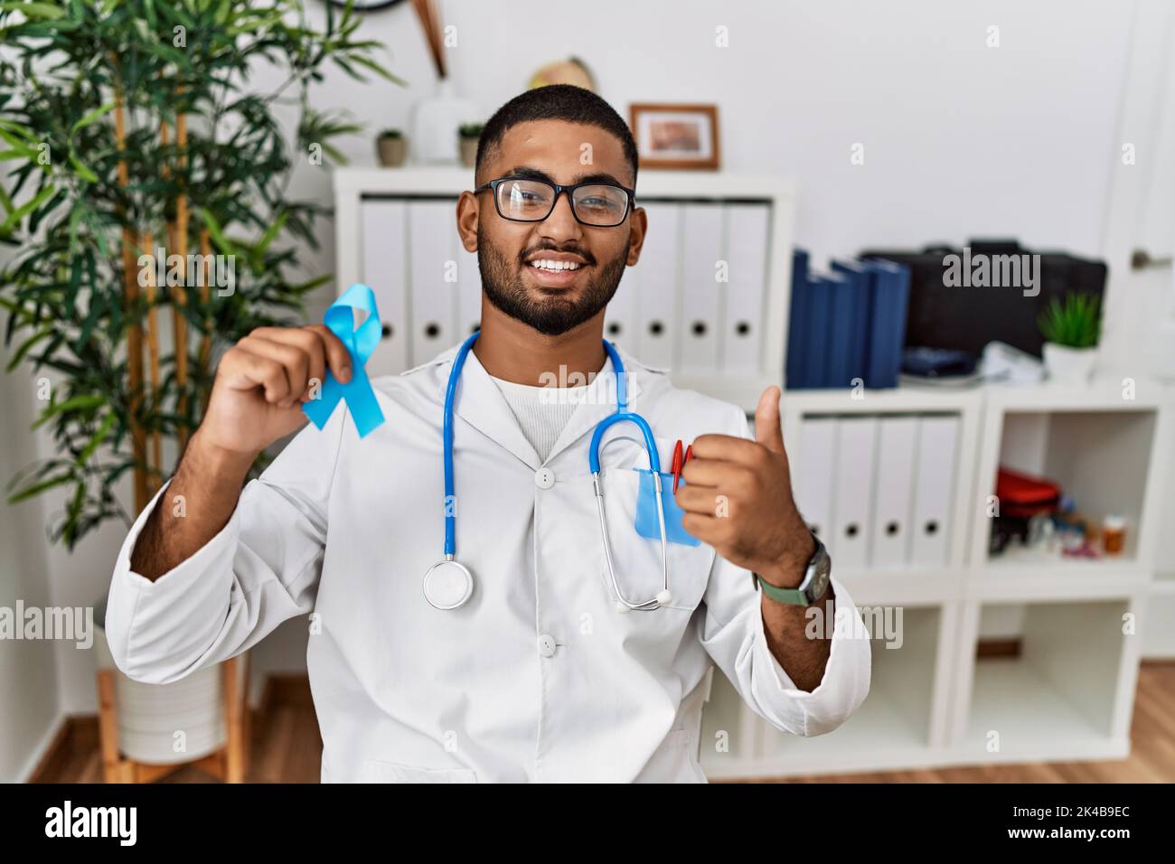 Young indian doctor holding blue ribbon smiling happy and positive ...