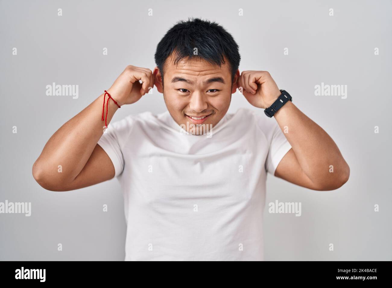 Young chinese man standing over white background smiling pulling ears ...