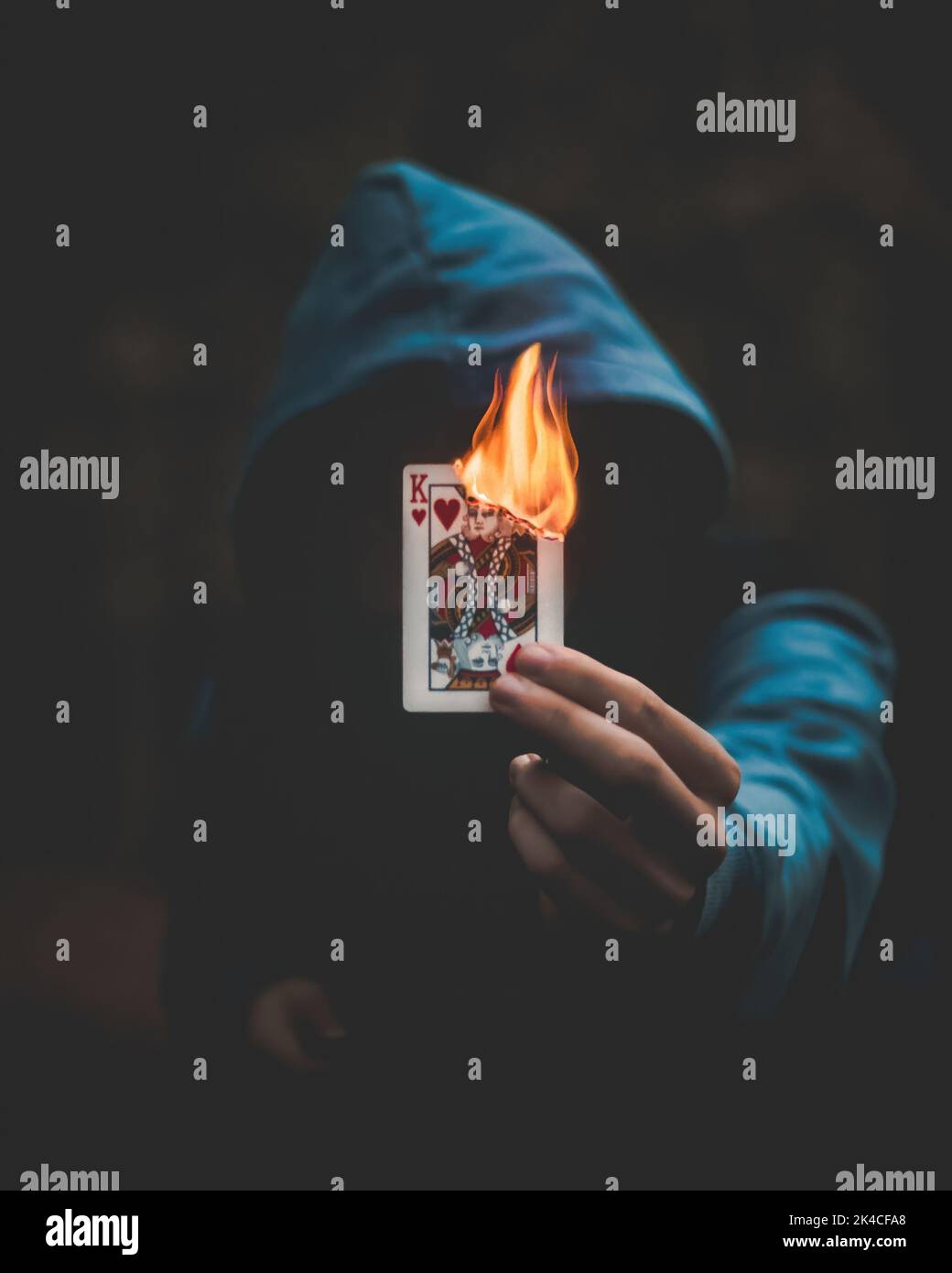 A vertical shot of a hooded male holding a burning king of hearts card ...