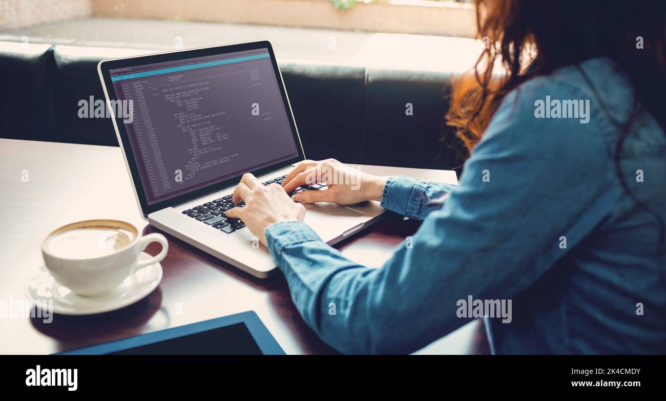 Caucasian female programmer sitting at desk with coffee, using laptop with coding on screen ...