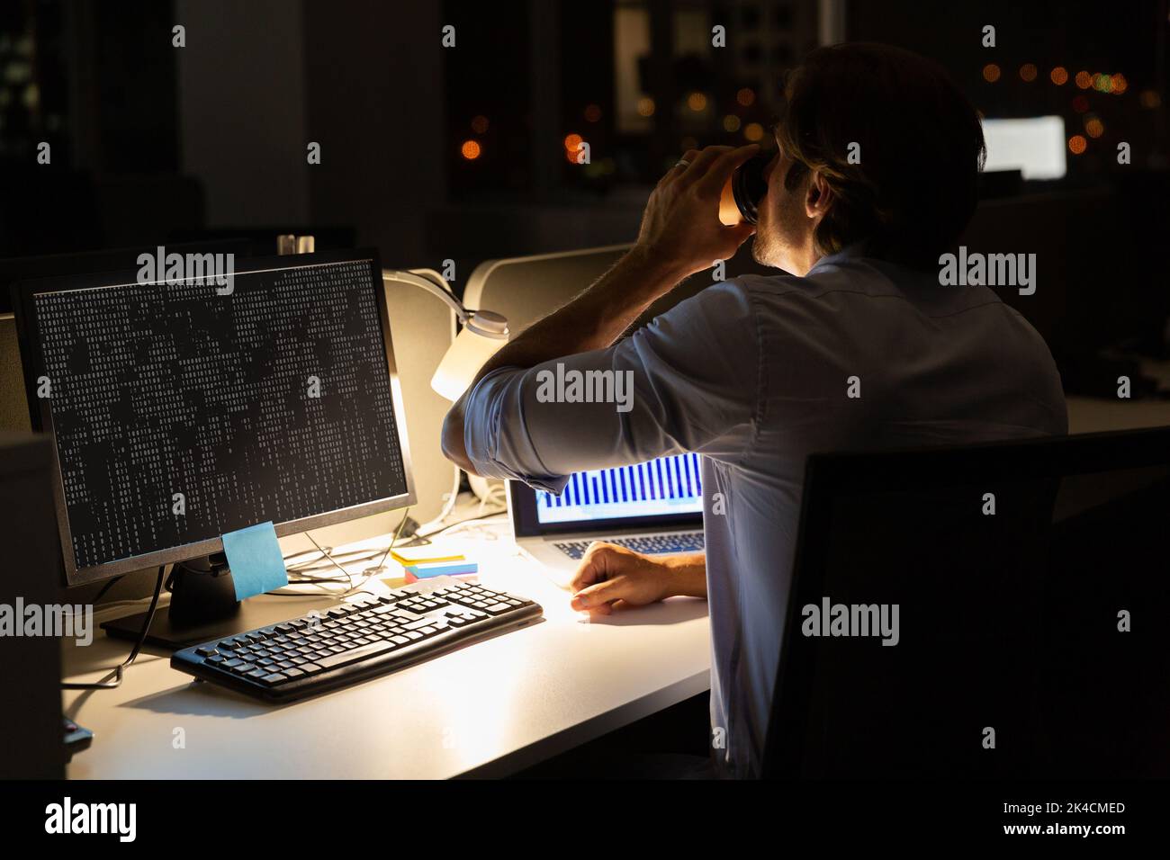 Caucasian Male Programmer Sitting At Desk Drinking Coffee Using Computer With Coding On Screen