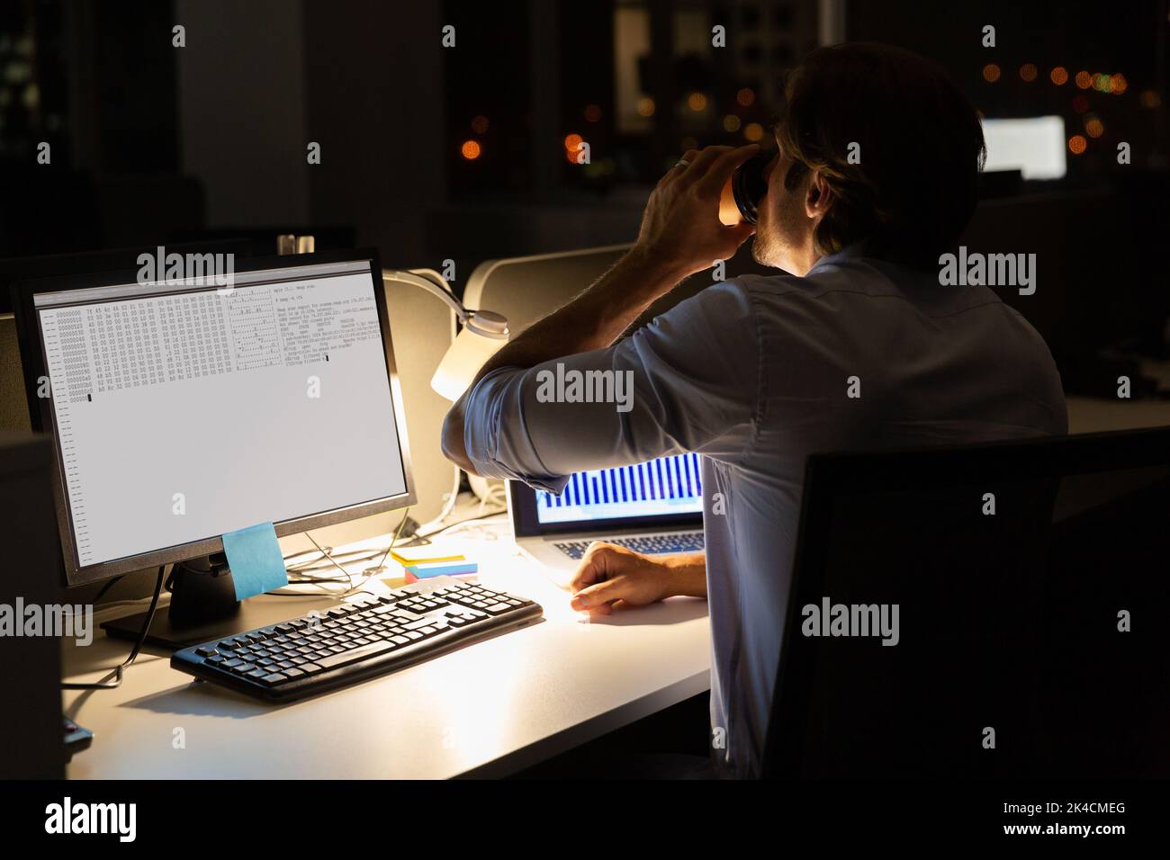 Caucasian Male Programmer Sitting At Desk Drinking Coffee Using Computer With Coding On Screen