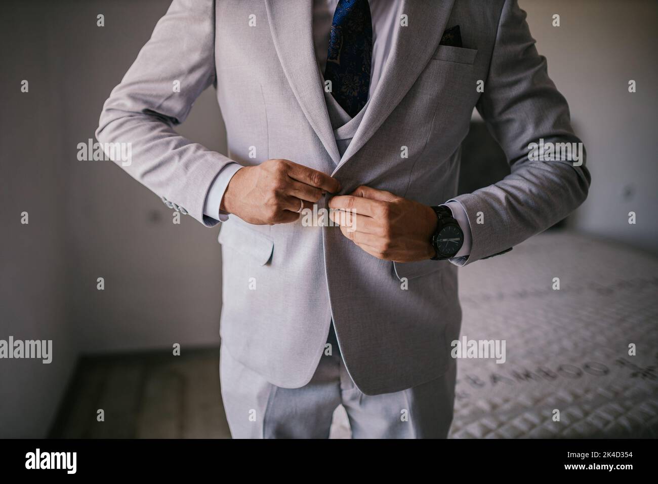Elegant man fastening a button on his light gray suit in the bedroom ...