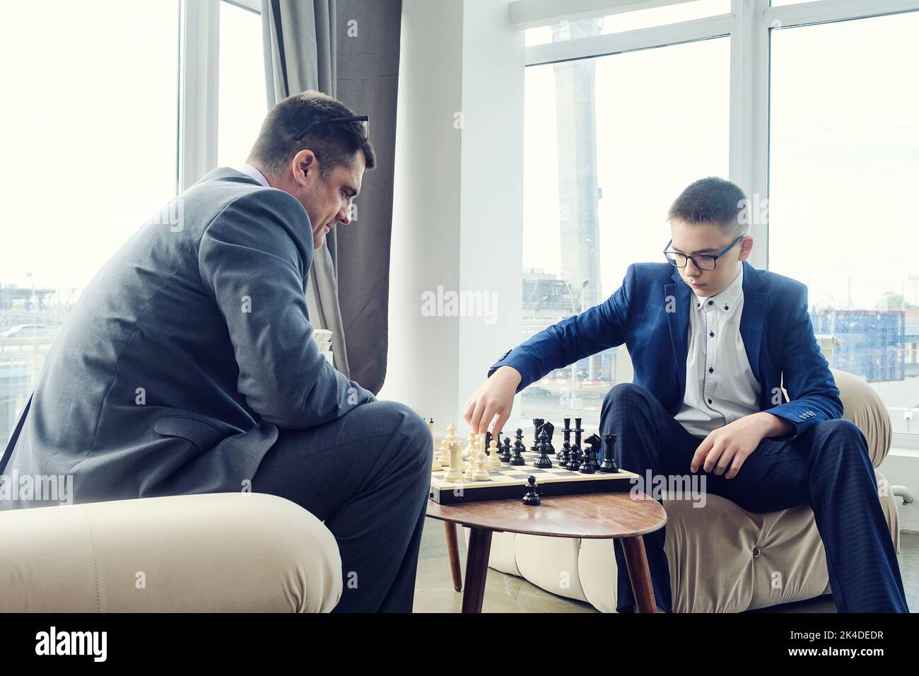 dad and son are sitting at a table by a wide window and playing chess ...