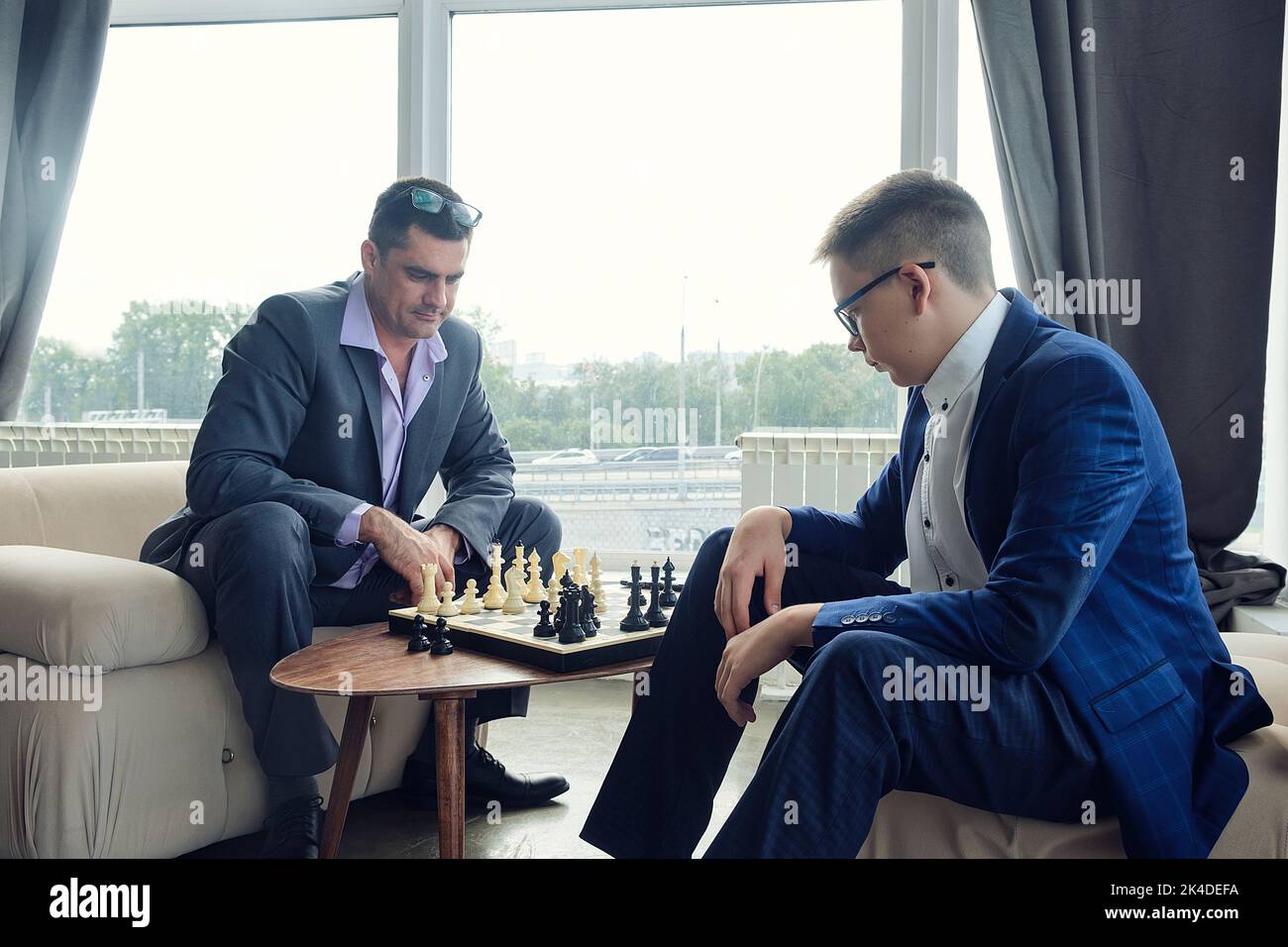 dad and son are sitting at a table by a wide window and playing chess ...