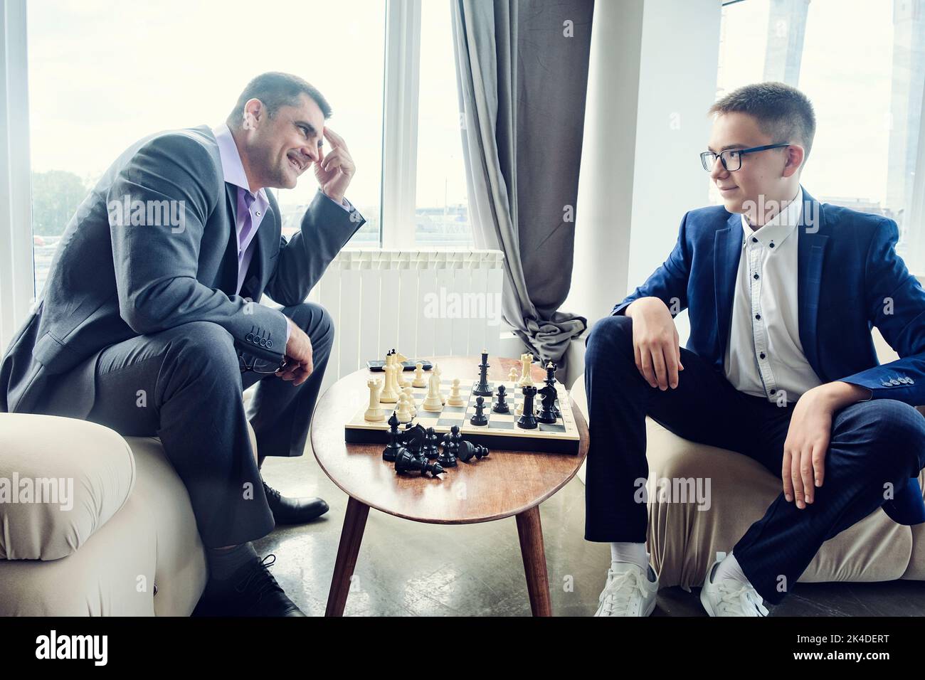dad and son are sitting at a table by a wide window and playing chess ...
