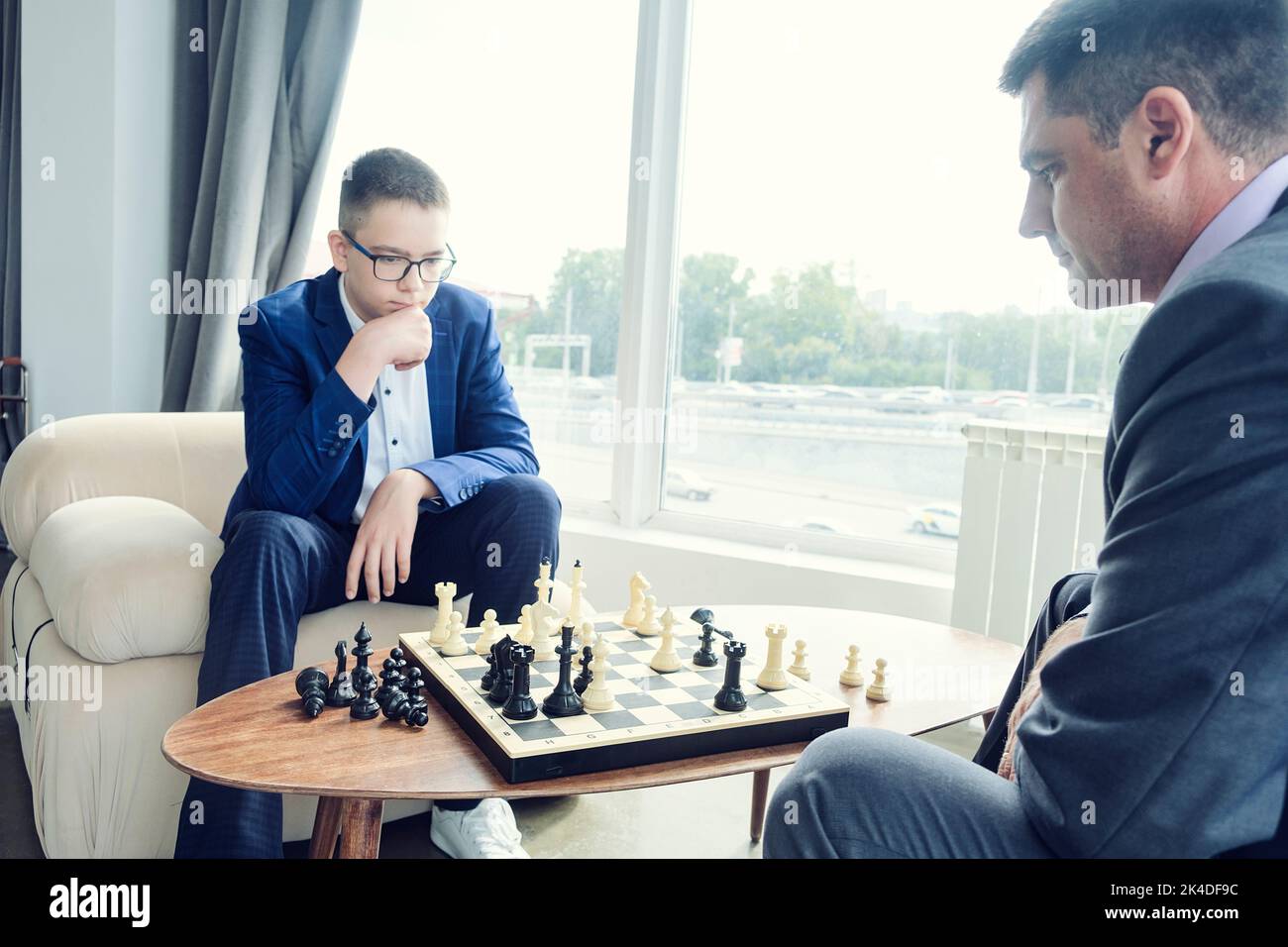 dad and son are sitting at a table by a wide window and playing chess ...