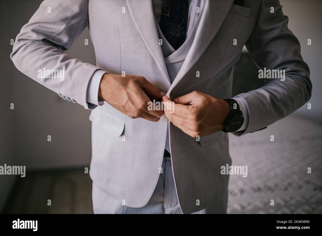 Elegant man fastening a button on his light gray suit in the bedroom ...