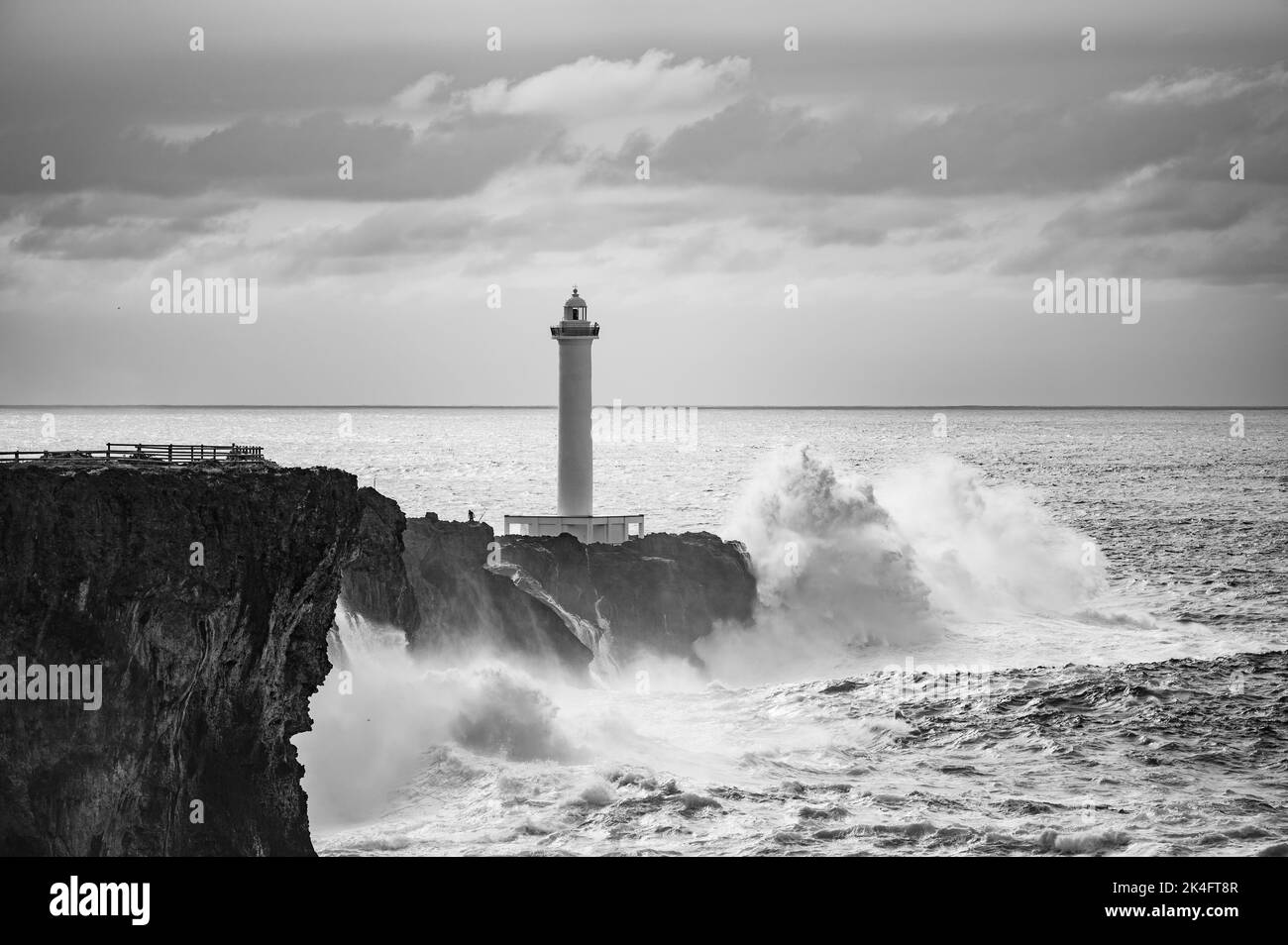 A greyscale of a lighthouse on the rock surrounded by stormy sea and ...