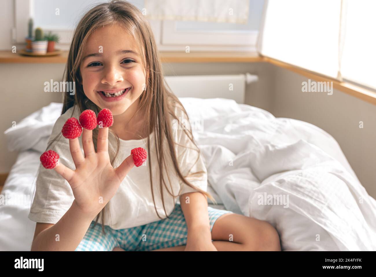 Funny little girl with raspberries on her fingers in bed in the morning ...