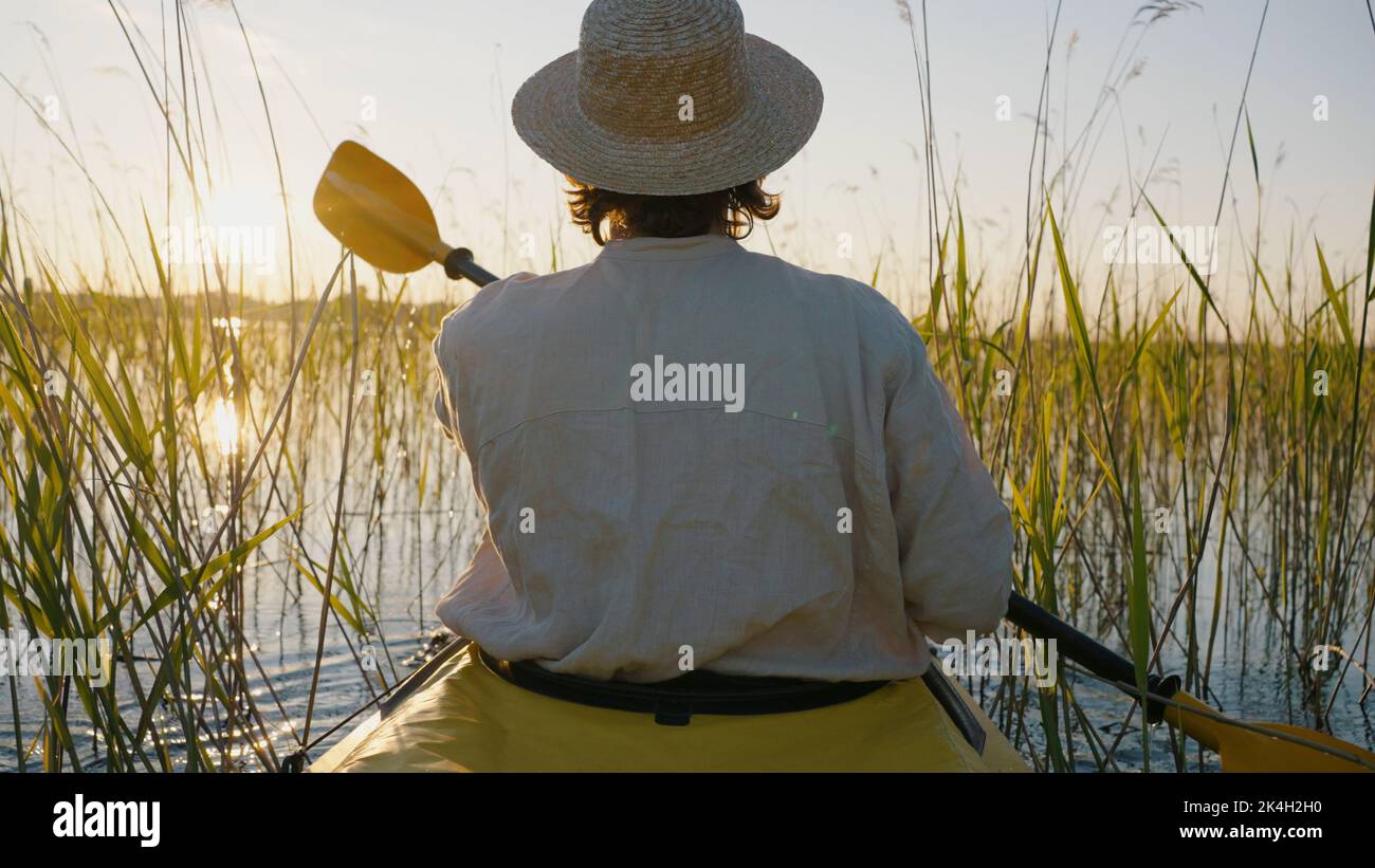 Person wearing straw hat sails on kayak using paddle through bulrush in ...