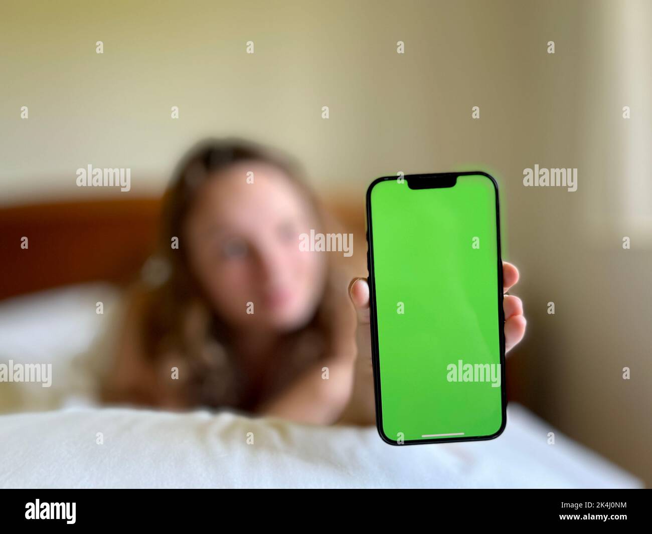 a girl in a white shirt a teenager lies on a white bed in her hands she holds a black iPhone 13 ...