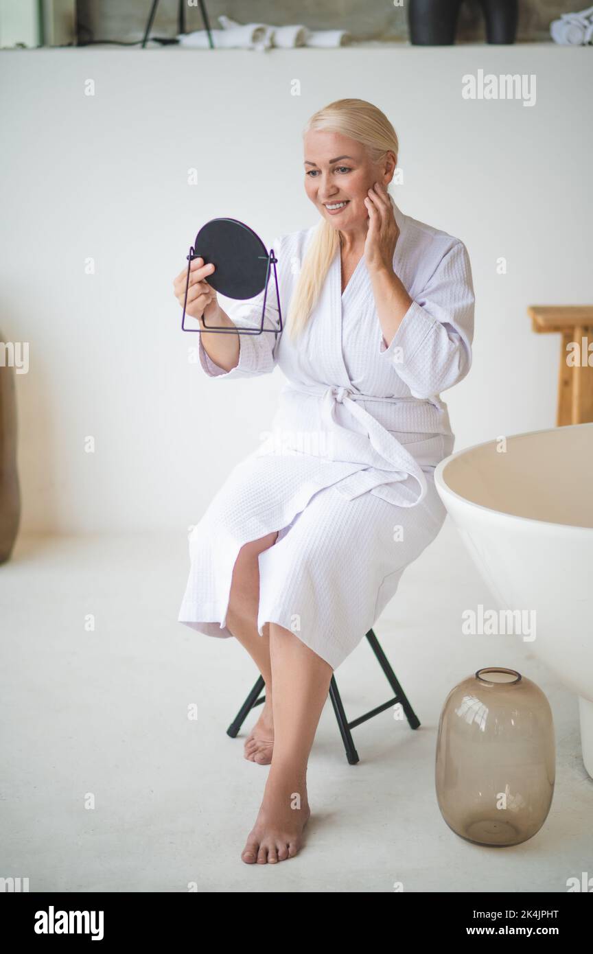 Joyous well-groomed female examining her skin in the handheld mirror ...