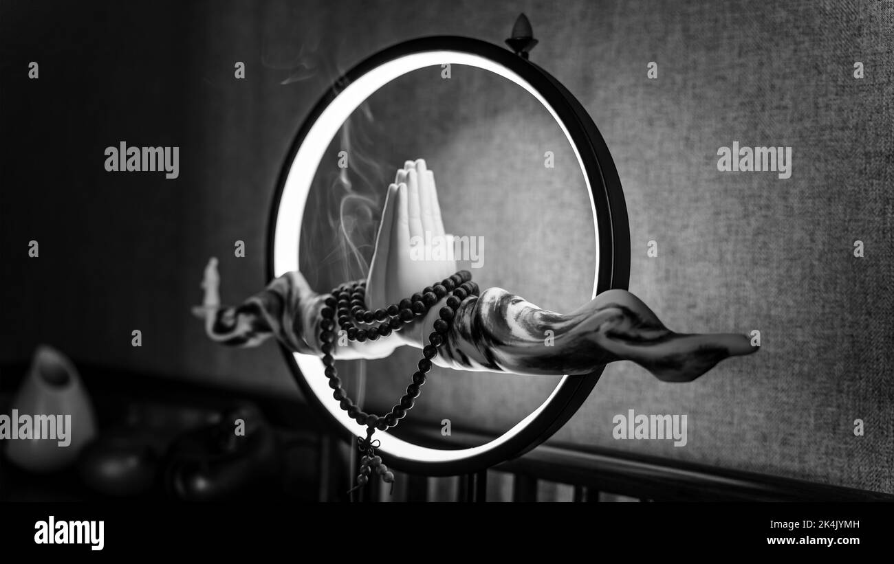 A grayscale closeup of a decorative lamp with hands in Namaste pose ...