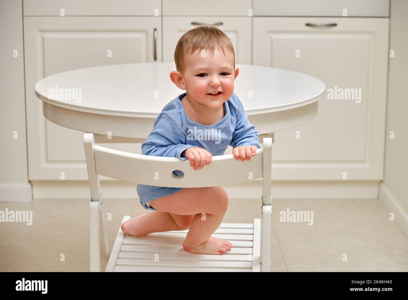 Toddler baby climbs onto a chair at the kitchen white table. A child in ...