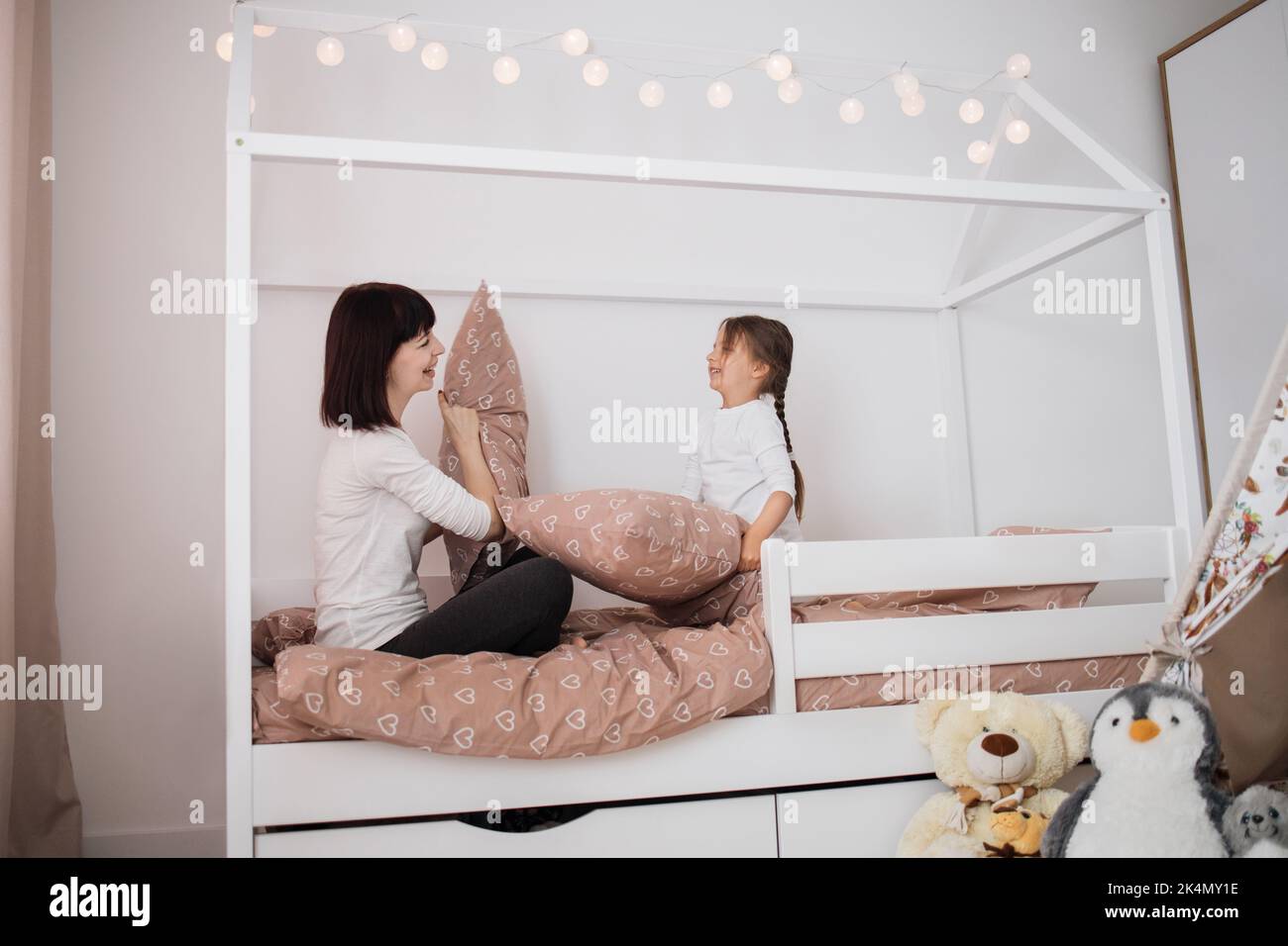 Overjoyed young mom and happy little daughter have fun engaged in pillow fight in bedroom ...