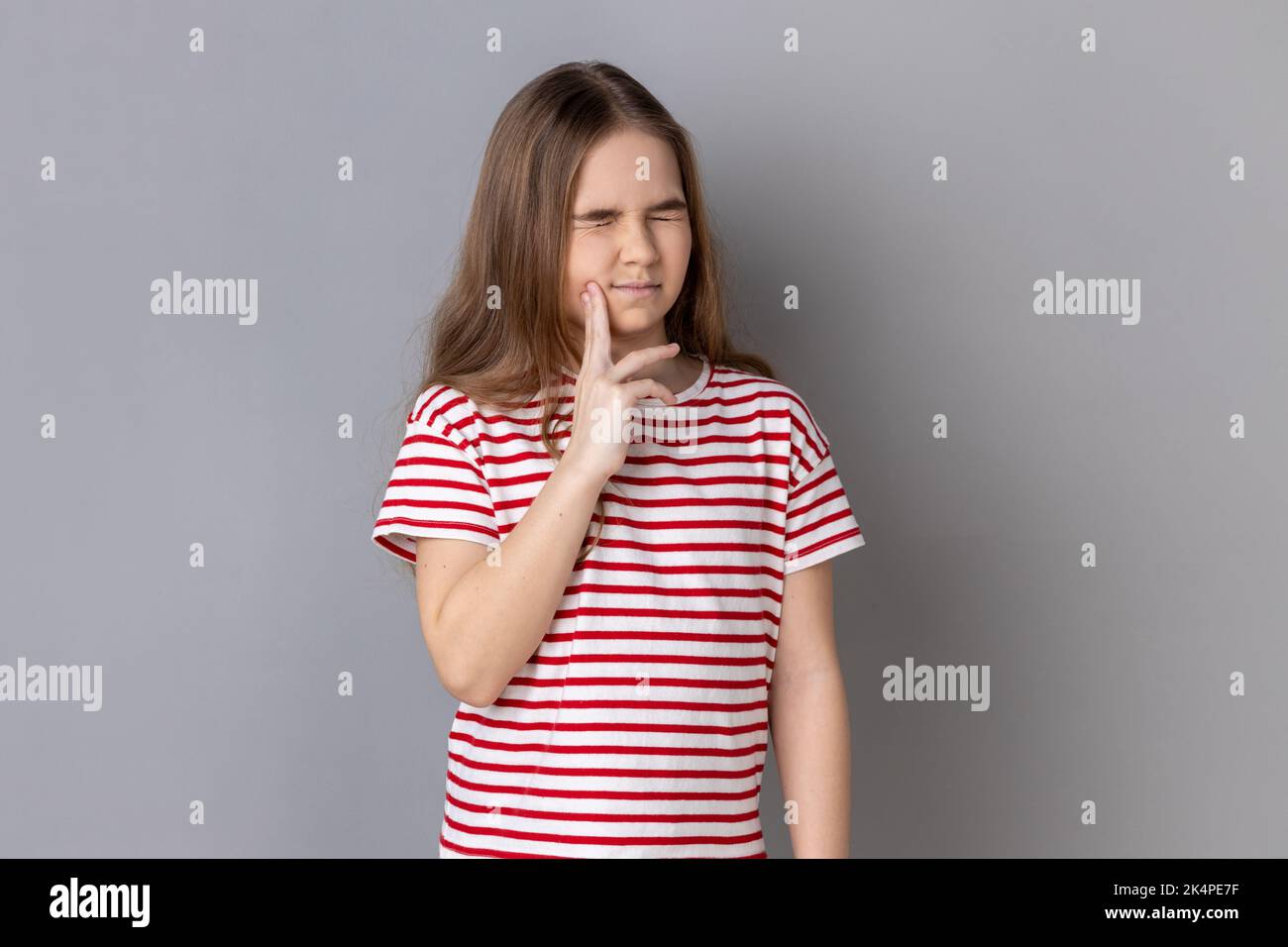 Sick little girl wearing striped T-shirt touching cheek, closing eyes ...