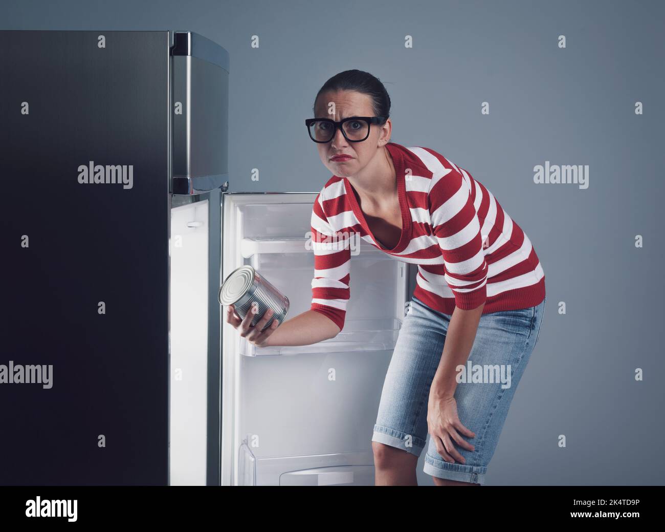 Sad hungry woman standing next to a open fridge, she is holding canned ...