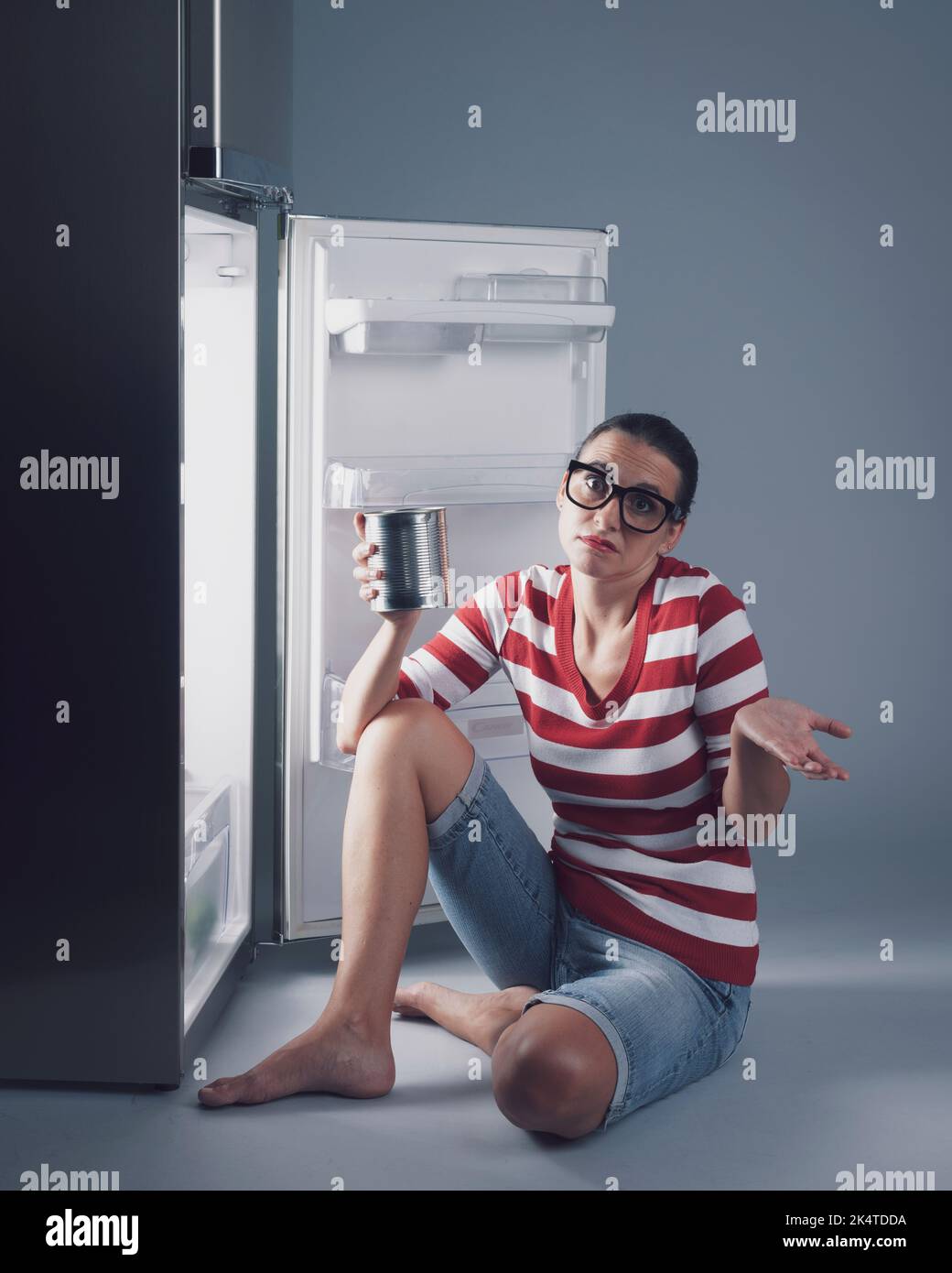 Sad hungry woman standing next to a open fridge, she is holding canned ...