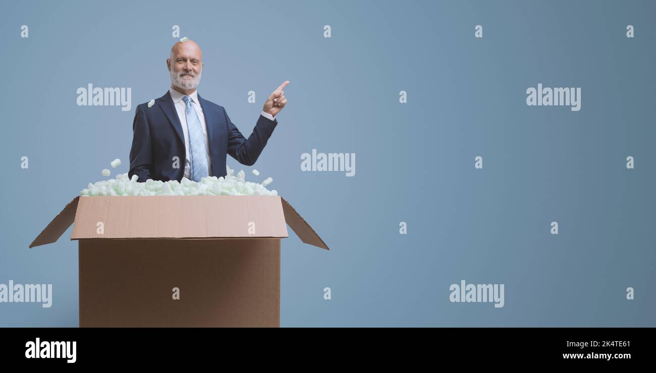 Confident smiling businessman coming out from a box and pointing finger ...