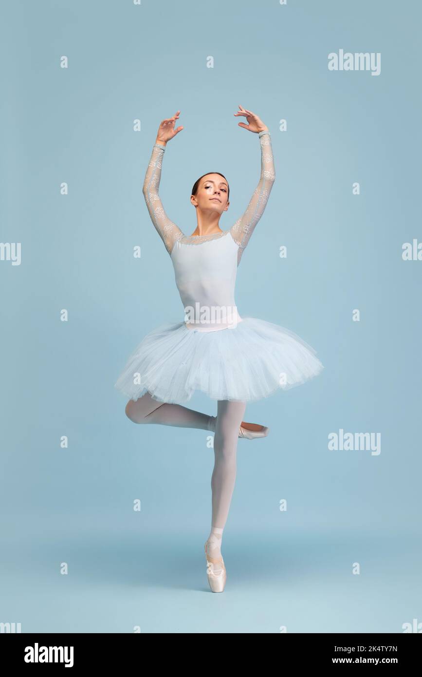Portrait of tender, beautiful, young ballerina dancing, performing isolated over blue studio ...