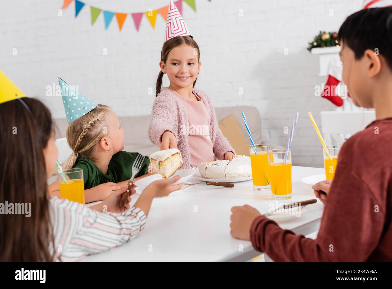 Cheerful child in party cap giving birthday cake to friend during ...