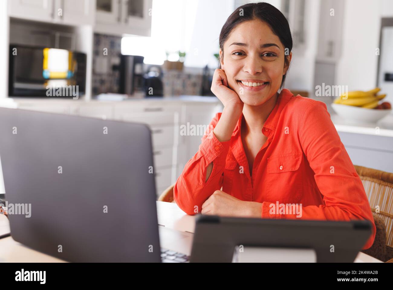 Portrait of happy biracial woman sitting at table in kitchen, using ...