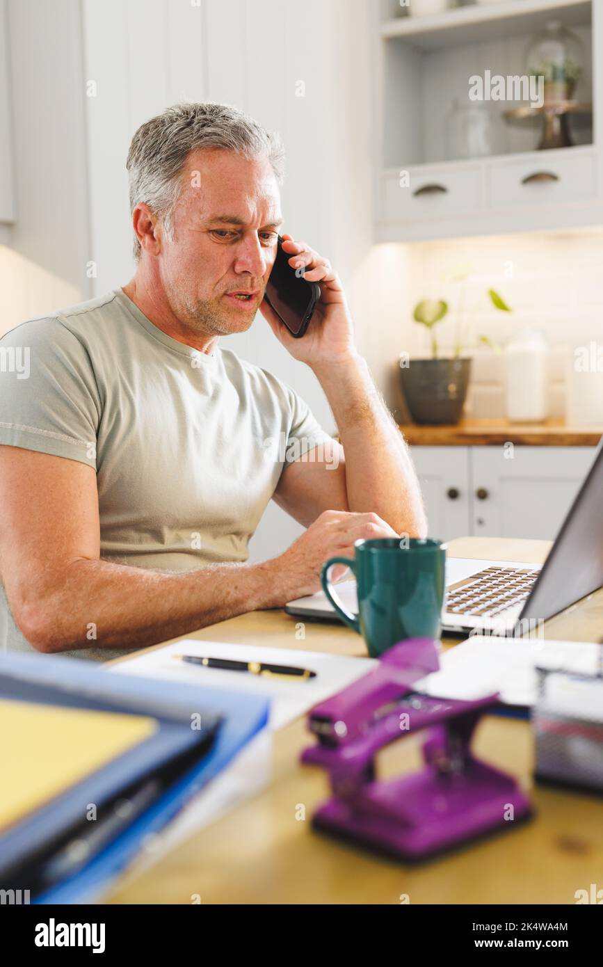 Happy caucasian man sitting at table in kitchen and using laptop and ...