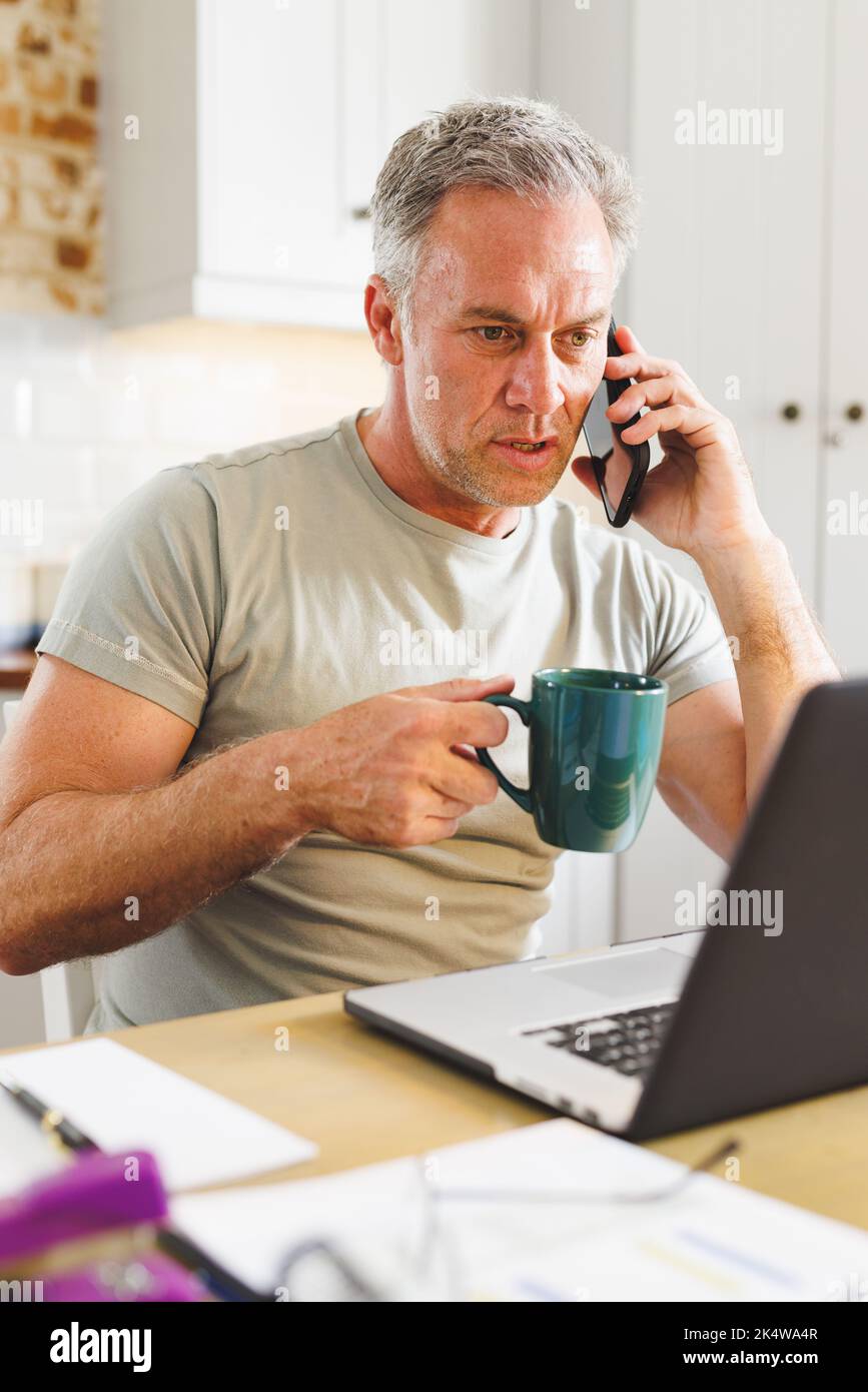 Happy caucasian man sitting at table in kitchen and using laptop and ...