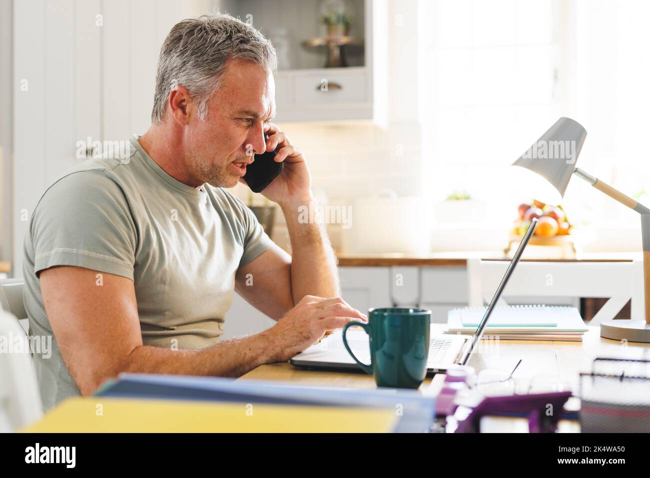 Happy caucasian man sitting at table in kitchen and using laptop and ...