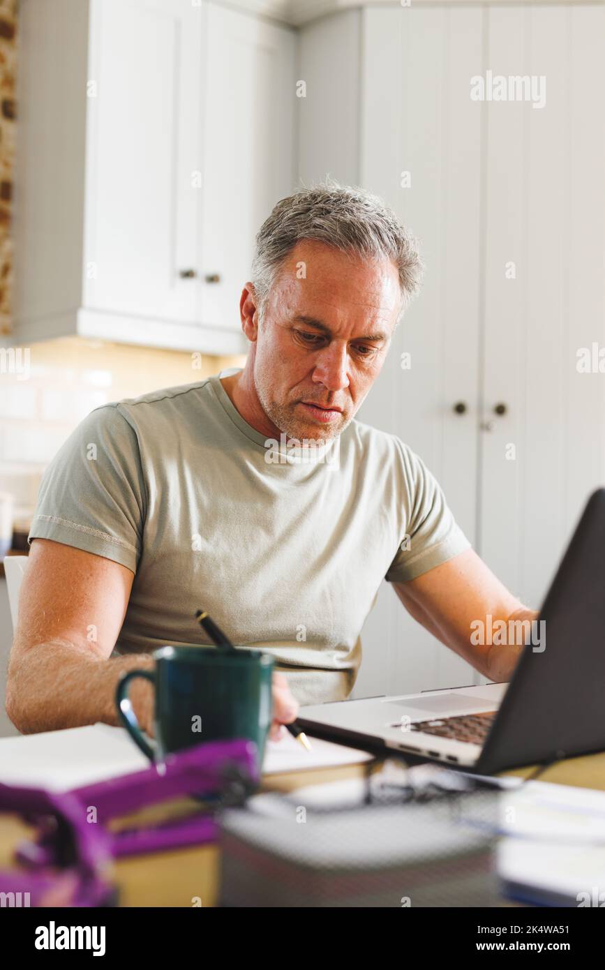 Happy caucasian man sitting at table in kitchen and using laptop and ...
