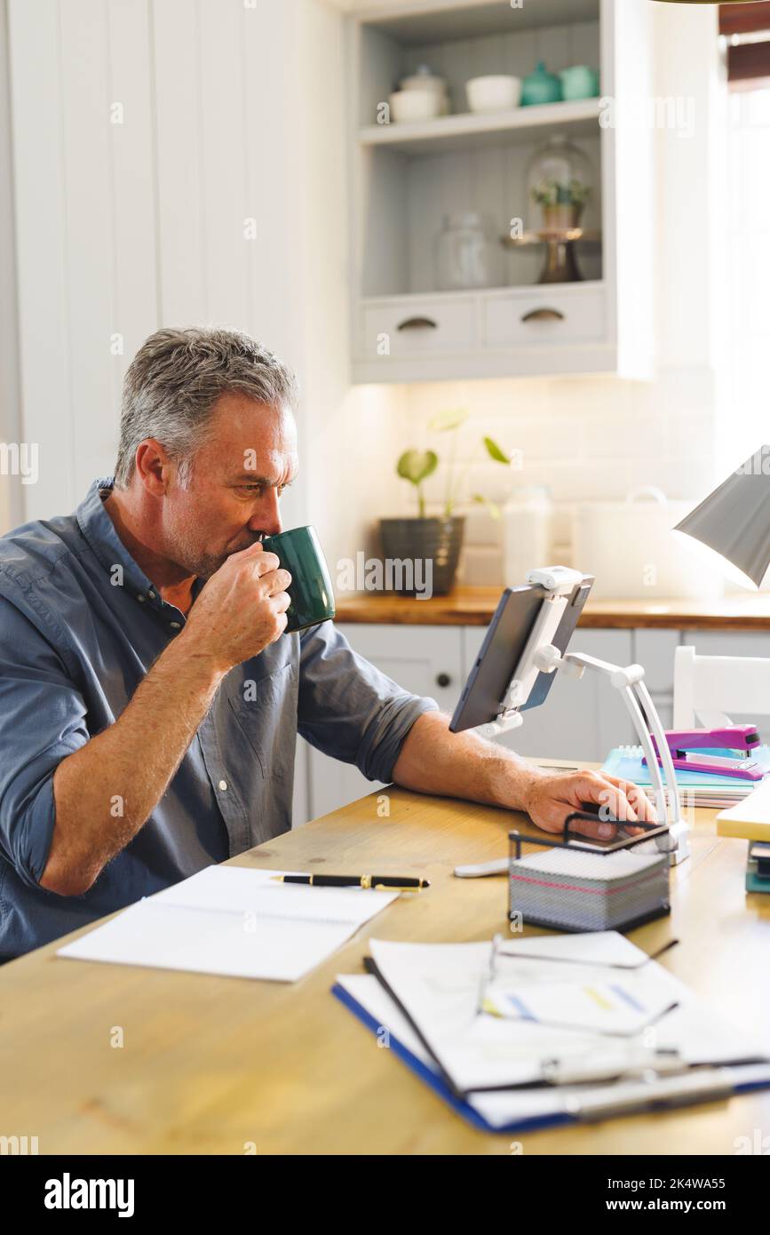 Happy caucasian man sitting at table in kitchen and using tablet and ...