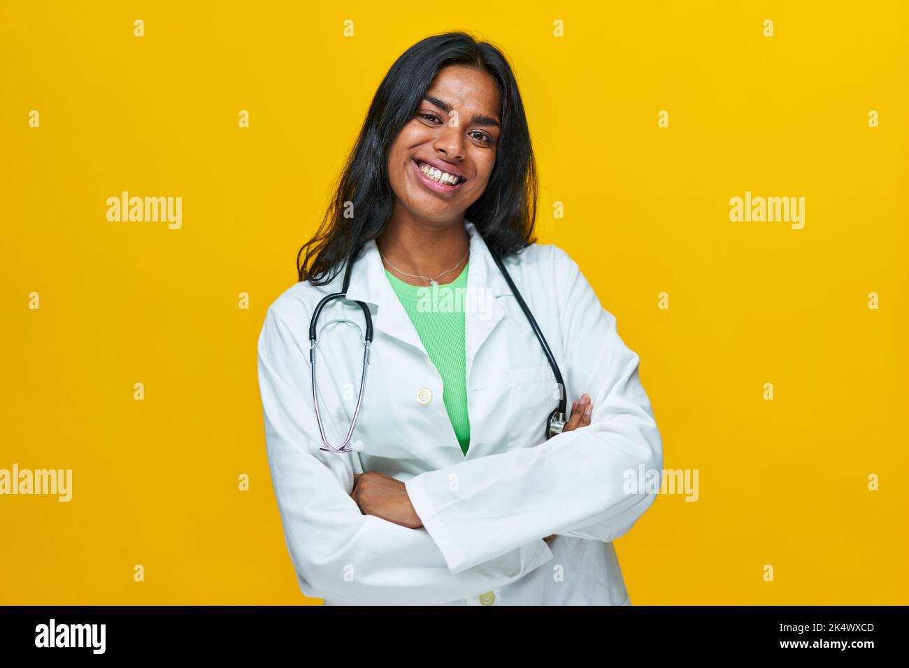 Cheerful young female doctor standing by a yellow background smiling ...