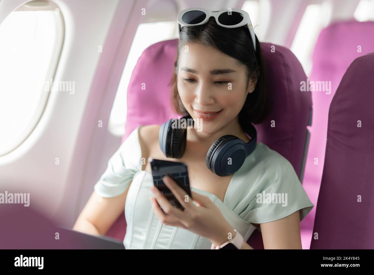 Joyful asian woman sits in the airplane and using mobile phone while go ...
