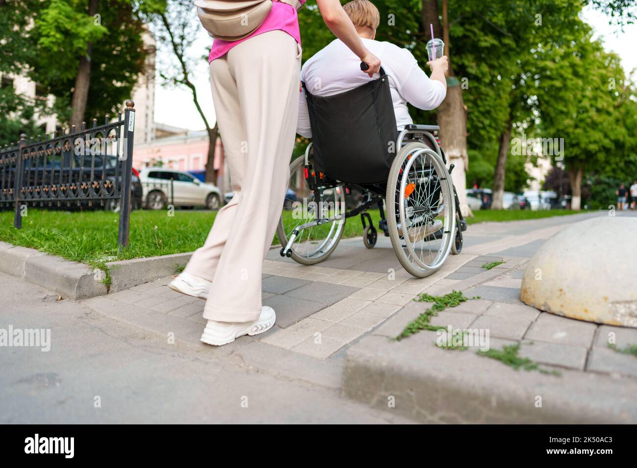 Back view of young woman helping mature woman in wheelchair in the city ...