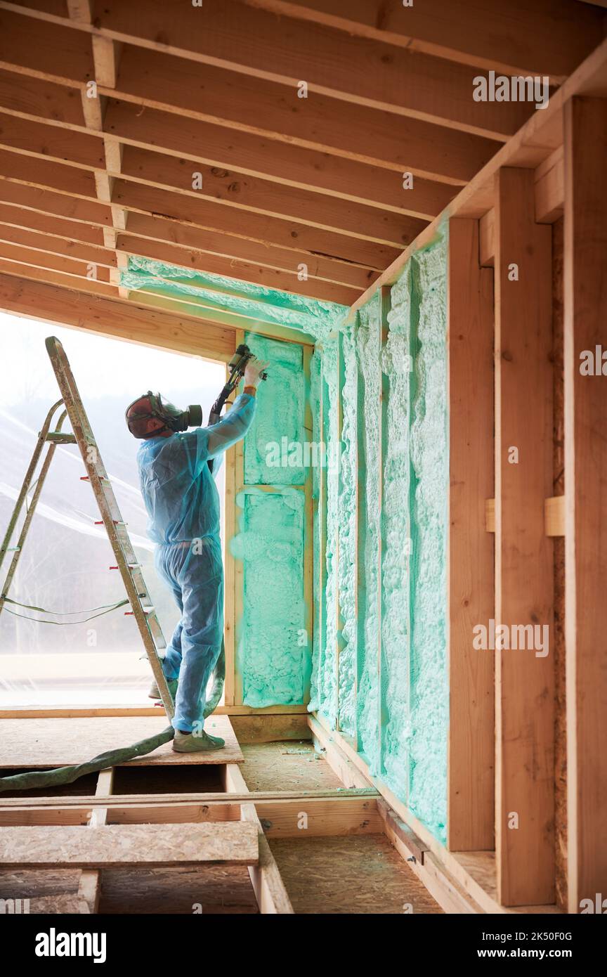 Male builder insulating wooden frame house. Man worker spraying ...