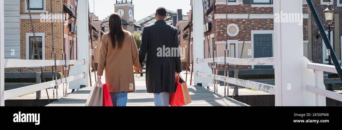 back view of trendy multiethnic couple with shopping bags holding hands ...
