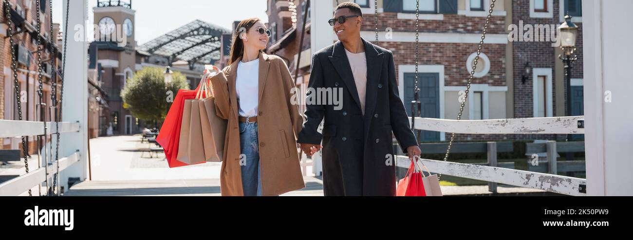 joyful multicultural couple in sunglasses carrying shopping bags and ...