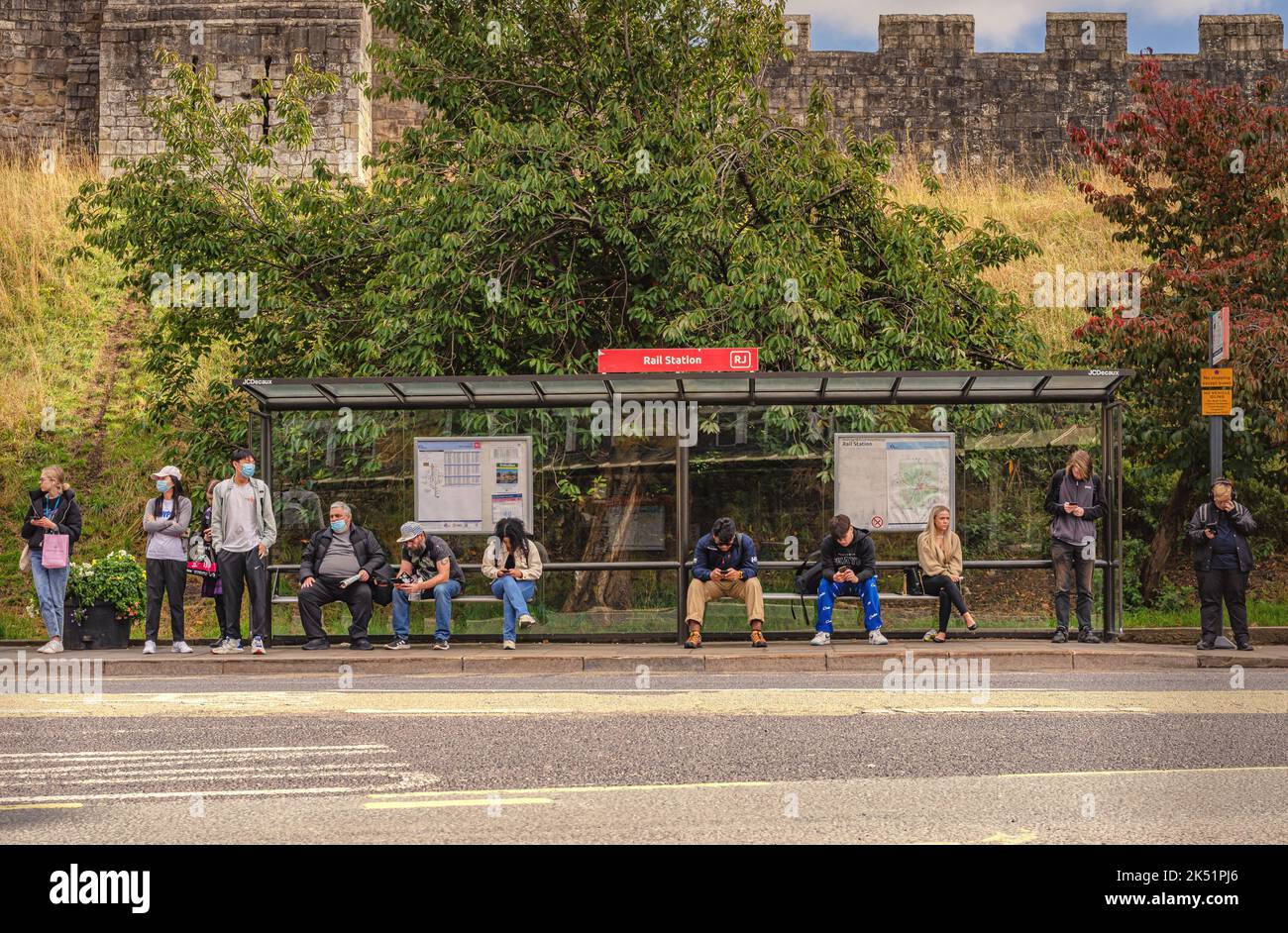 Passengers wait for a bus in a line by a bus shelter. Some are sitting ...