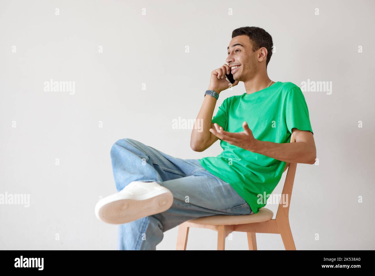 Excited arab guy speaking on cellphone, sitting on chair over white ...