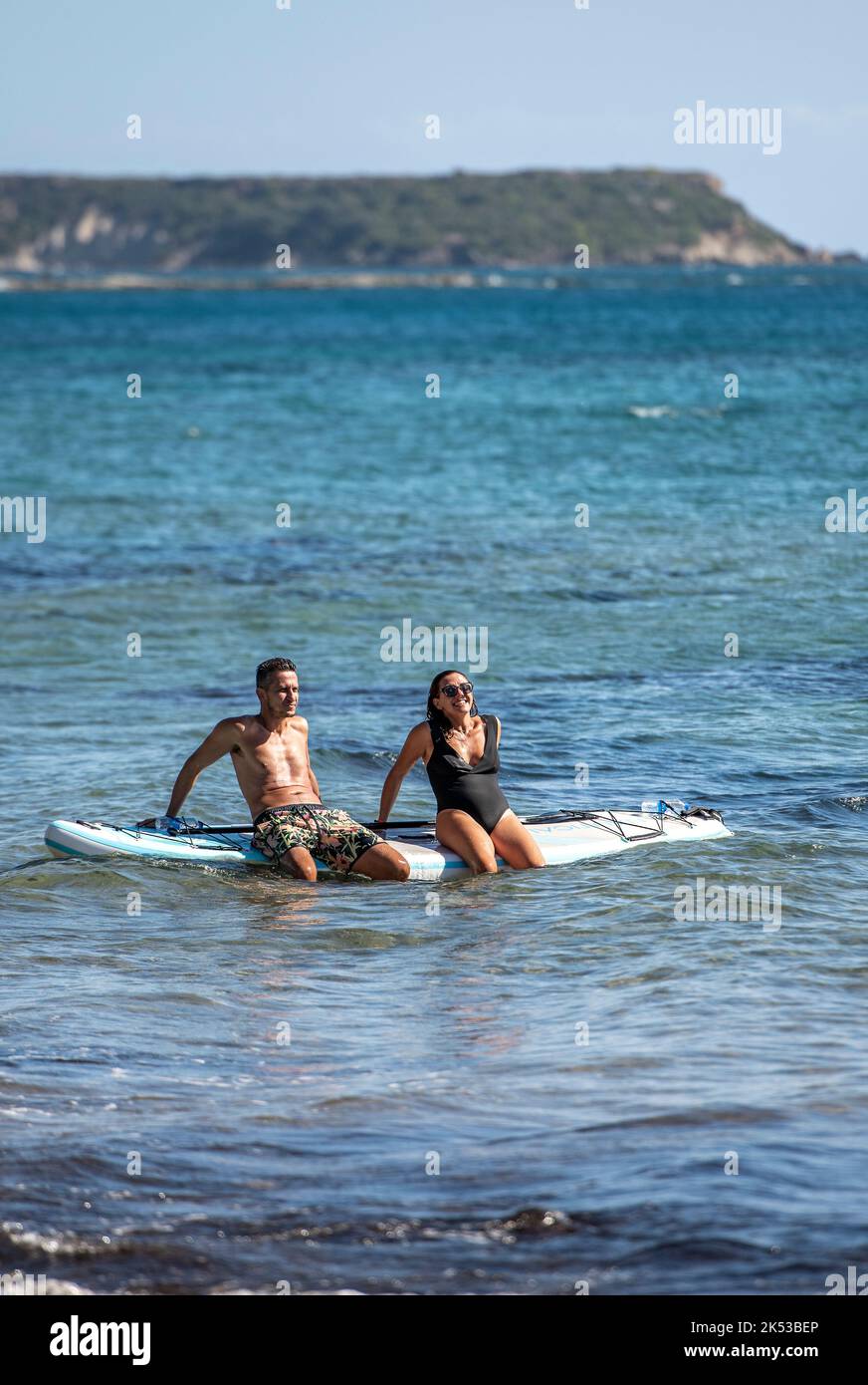 Young Couple Sitting On A Surfboard In The Summer Sunshine On A Beach On The Greek Island Of