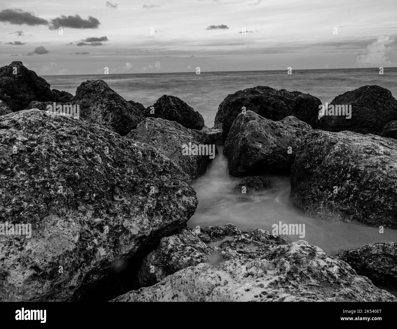 A grayscale of big mossy rocks in the sea with a horizon in the ...