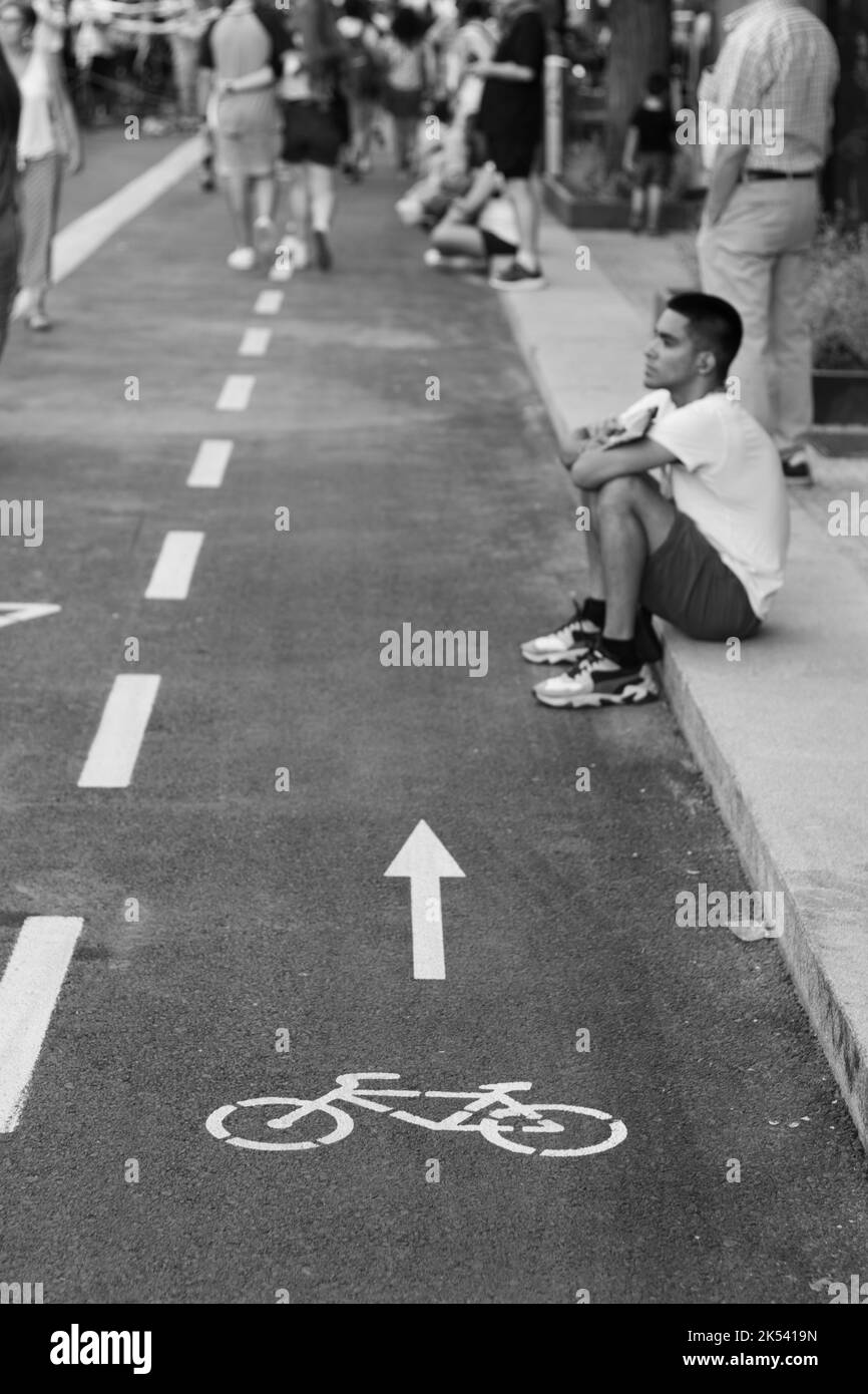 A grayscale of a male sitting on the sidewalk curb on the street with ...